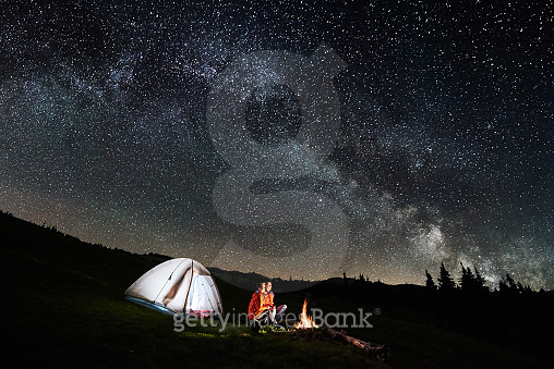 Night camping. Couple tourists sitting at a campfire near illuminated ...