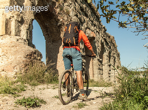 Cyclist riding bike on dirt road under ancient roman aqueduct in a park ...