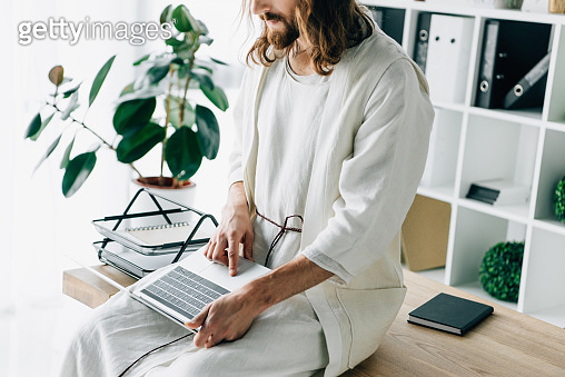 partial view of Jesus sitting on table and working on laptop in modern ...