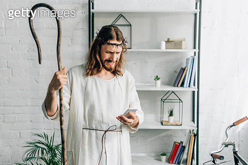 emotional Jesus in robe with wooden staff using smartphone at home ...