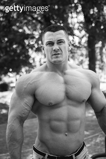 A male bodybuilder stands with bare torso in denim shorts in the park ...