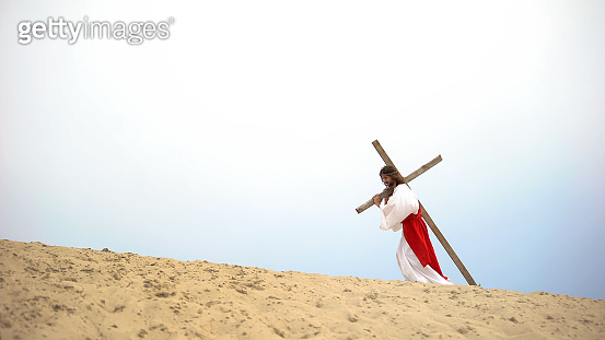 Personification of Jesus Christ carrying heavy cross in desert ...