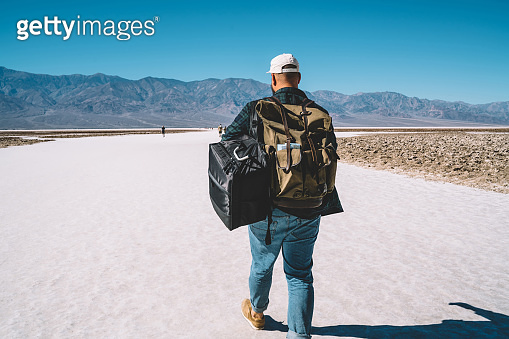 Young man wanderlust with big touristic backpack strolling on dry surface of death walley ...