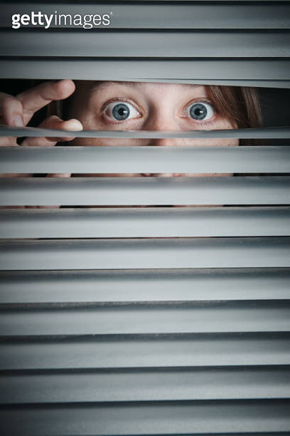 Startled young woman with wide blue eyes looks through blinds ...