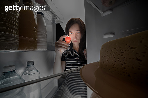 Sad girl looking into fridge and holding single tomatoes in her hand ...