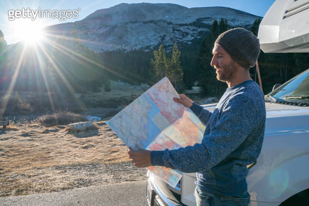 Road trip concept; man sitting on vehicle's hood checking at road map ...