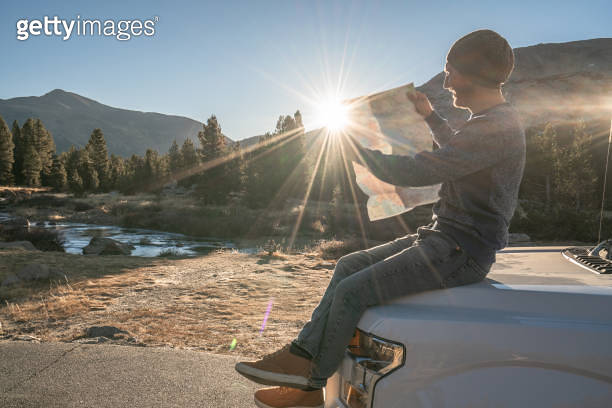 Road trip concept; man sitting on vehicle's hood checking at road map ...