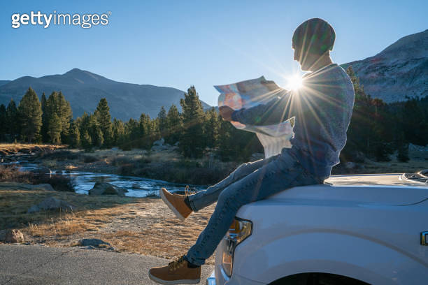 Road trip concept; man sitting on vehicle's hood checking at road map ...