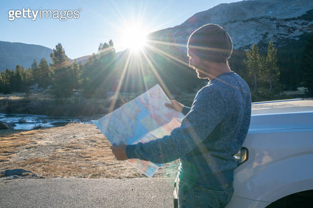 Road trip concept; man sitting on vehicle's hood checking at road map ...