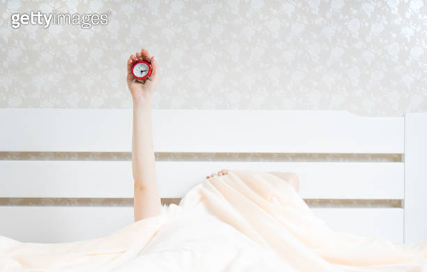 Hands of a woman with alarm clock from under the blanket. Early morning ...
