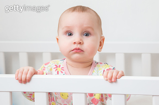 Seriously, confused baby girl in colorful pajama in white crib. Front view. Closeup. 이미지 ...