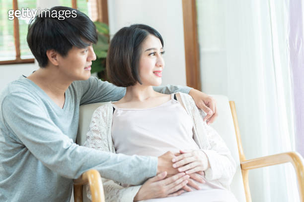 Cheerful expectant mom and dad sitting on couch, look out window ...