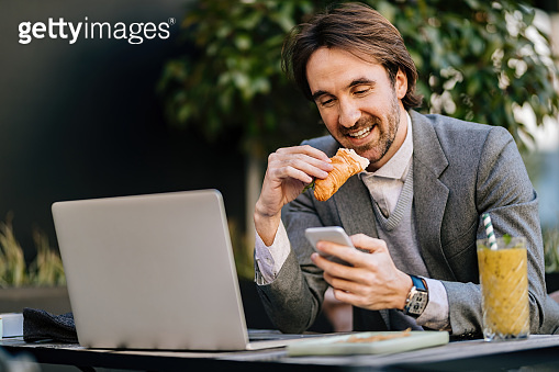 Happy businessman using cell phone while eating croissant in a cafe ...