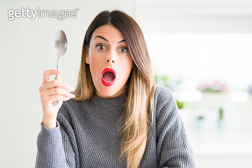 Young beautiful woman holding silver spoon at home scared in shock with ...