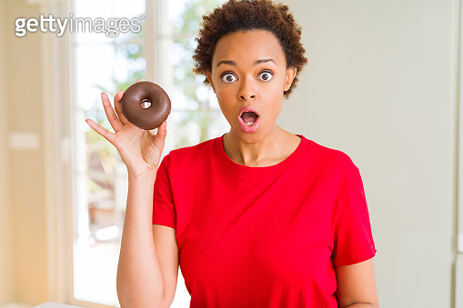 Young african american woman eating chocolate donut scared in shock ...