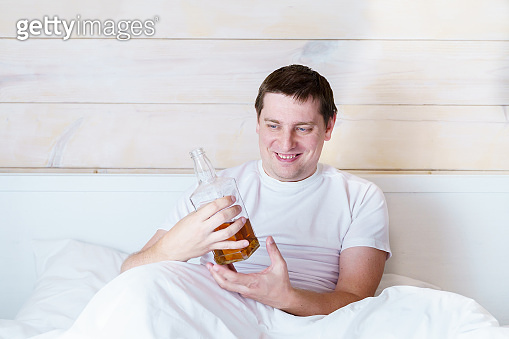 Drunk rowdy man with angry face, bottle of alcohol sits drinking in bed ...