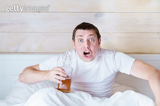 Drunk rowdy man with angry face, bottle of alcohol sits drinking in bed ...