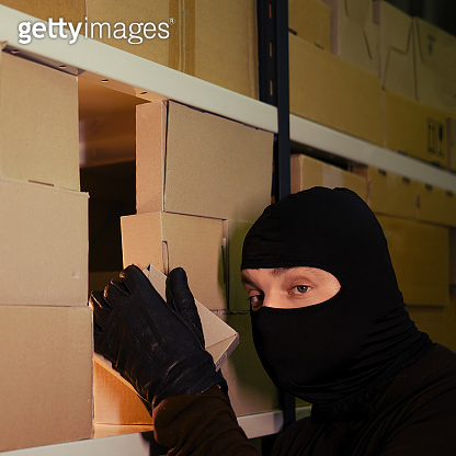 Thief in a black ski mask steals a box with a parcel in a warehouse at ...