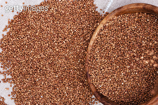 Raw roasted buckwheats from round wooden bowl pour on cloth surface ...