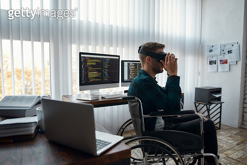 Young male web developer in a wheelchair wearing virtual reality glasses while working at his ...