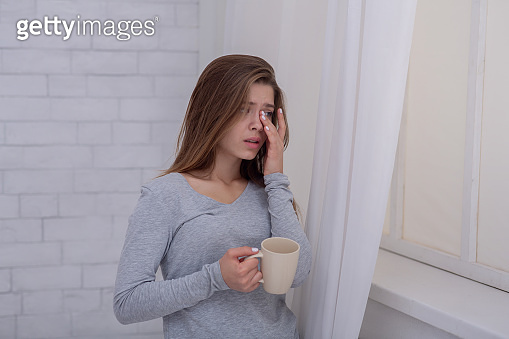Unhappy young lady standing near window with cup of coffee and crying ...