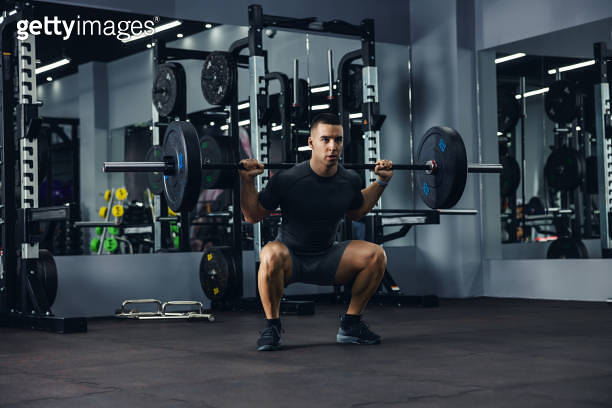 A side portrait of a bodybuilder in grey doing squats using a barbell ...