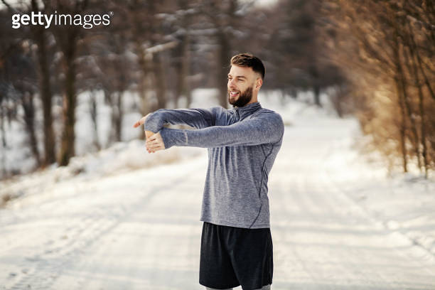 Fit sportsman doing warm up exercises while standing in nature at snowy ...