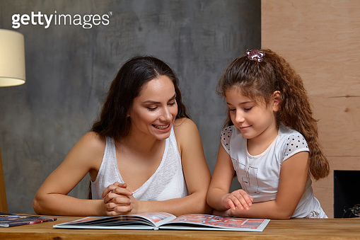 Portrait of a mother helping her small sweet and cute daughter to make her homework indoors ...