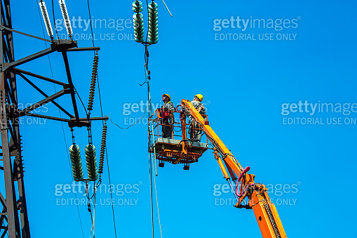 High voltage power line transmission tower workers with crane and blue ...