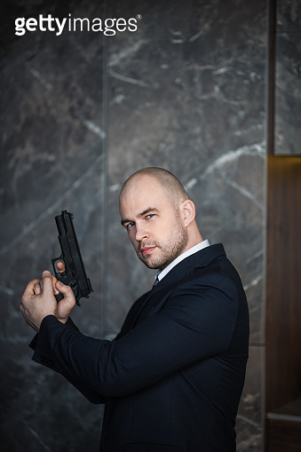 A bald man in a suit with a gun is standing against the wall in a dark ...