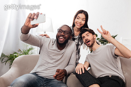 Portrait of three smiling friends sitting on chairs at home and taking ...