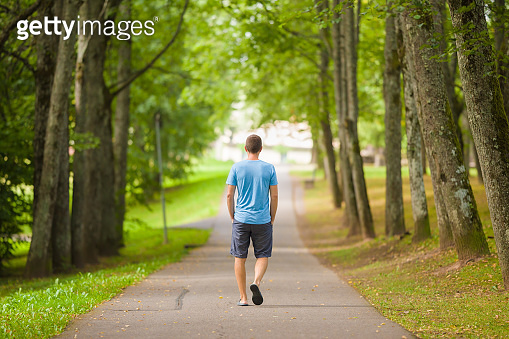 Young adult man slowly walking through alley of green trees on sidewalk ...