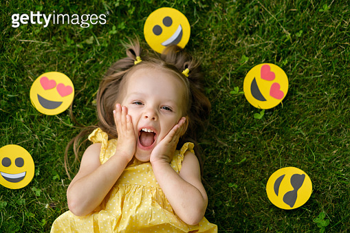 A funny little girl surrounded by a variety of paper emoticons looks up ...