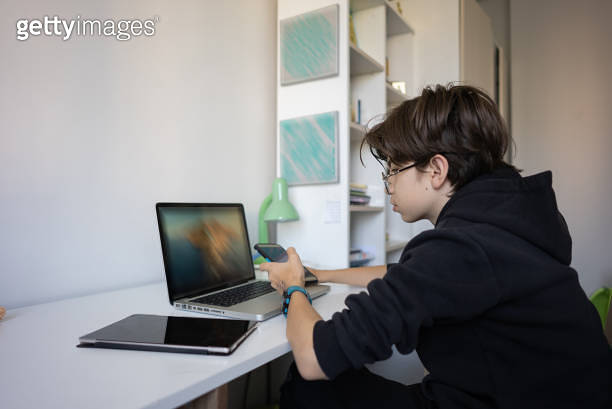 Boy using phone and laptop while doing homework 이미지 (1392369456) - 게티이미지뱅크