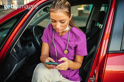 A smiling young nurse dressed in her scrubs uniform sitting in her car ...