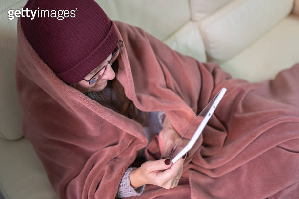 Woman wrapped in a blanket and wearing a wool hat looking at a tablet ...