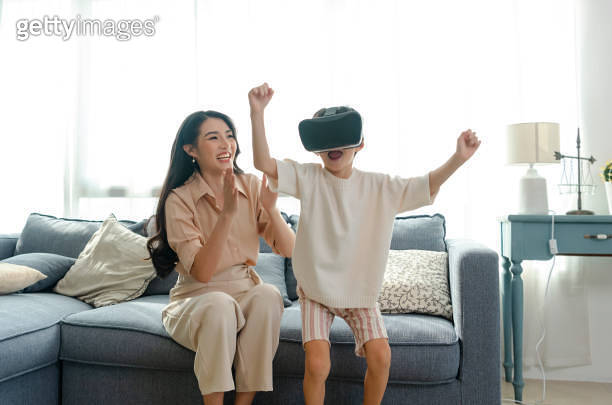 Smiling mother looking son playing games using virtual reality headset ...