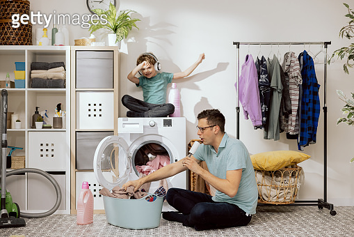 Baby sits on the washing machine while dad puts the laundry in. The ...