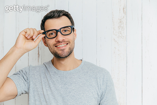 Attractive male model with stubble, looks joyfully through spectacles ...