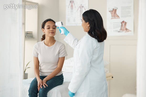 Shot of a young woman sitting in the clinic and getting her temperature checked by her doctor ...