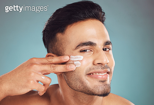 Portrait of one smiling young indian man applying moisturiser lotion to his face while grooming ...