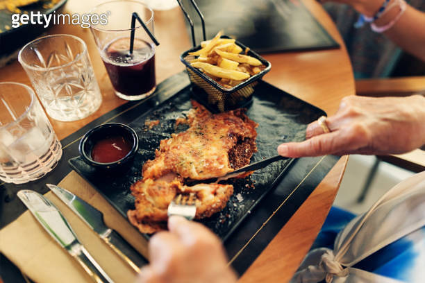 Senior woman cutting fresh fried cutlet with French fries and tomato ...