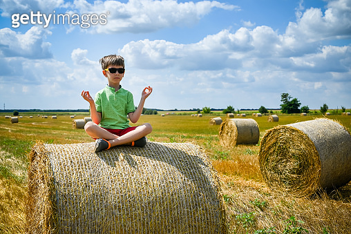 Little boy sits on a round haystack relaxed and meditating. Field with ...