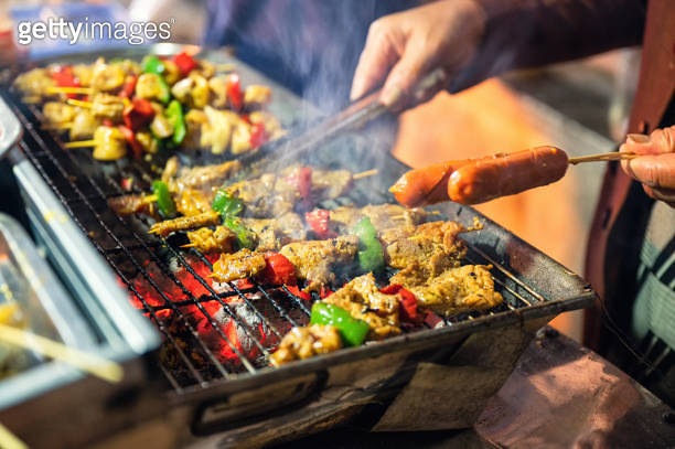 Seller using tongs grilling barbeque pork and sausage on charcoal stove ...