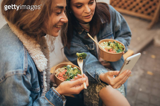 Two young women female friends using smartphone sitting outdoors eating ...