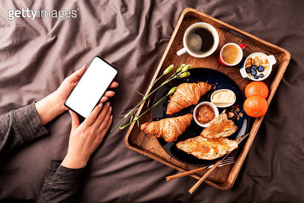 Woman using smartphone while eating continental breakfast in bed. Blank ...