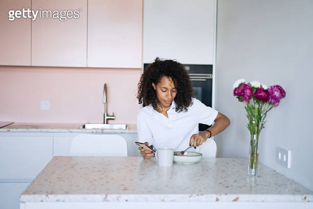 Woman sitting at kitchen counter having breakfast and typing a text ...