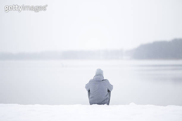Young adult man sitting alone on snow at lake shore and looking far ...