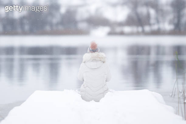 Young adult woman sitting on snow covered edge of footbridge at lake ...
