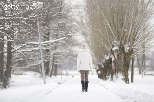 Young adult woman slowly walking on white snow covered sidewalk at park ...